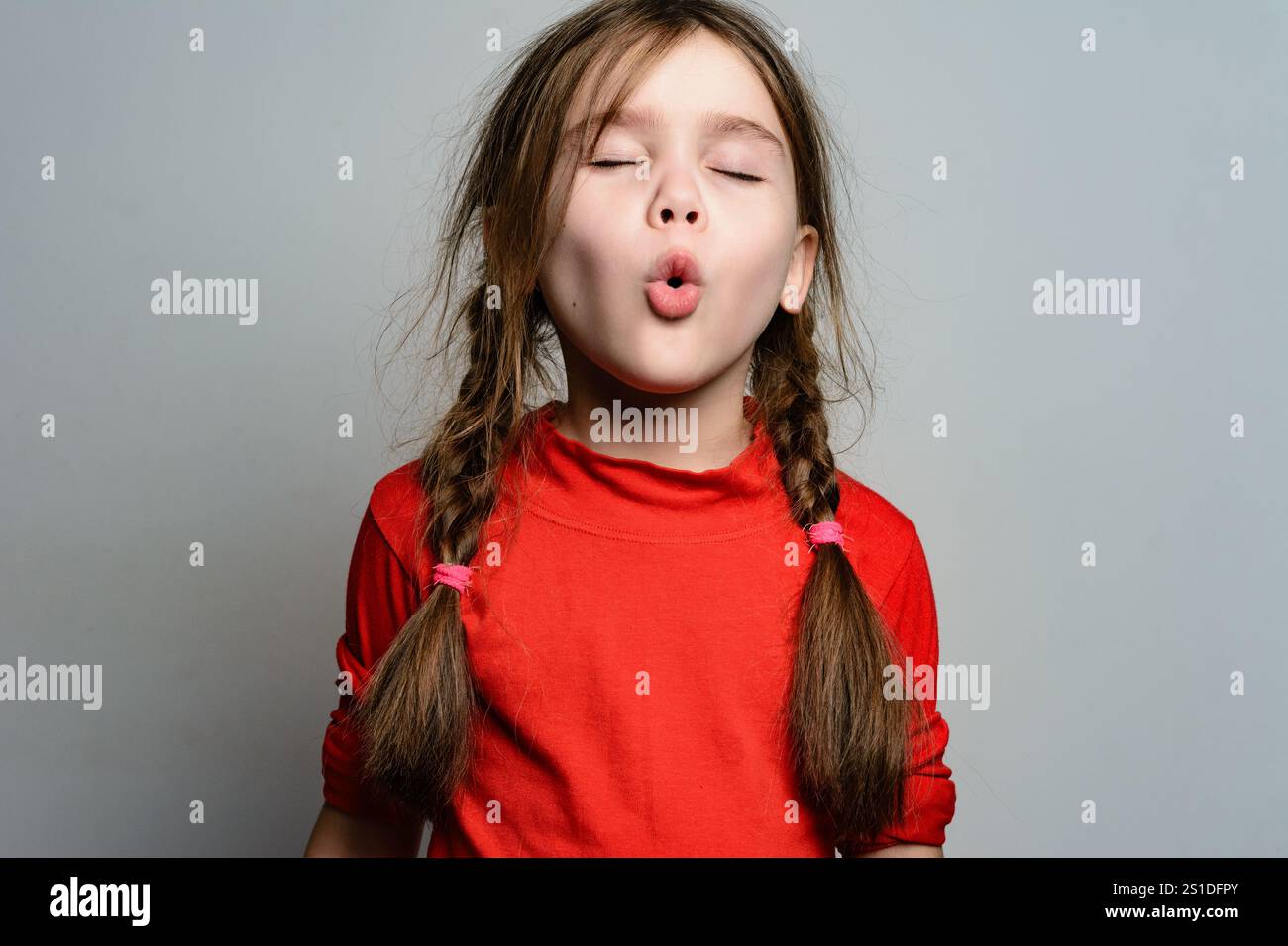 A young girl with brown pigtails in a red shirt makes a funny face. Her ...