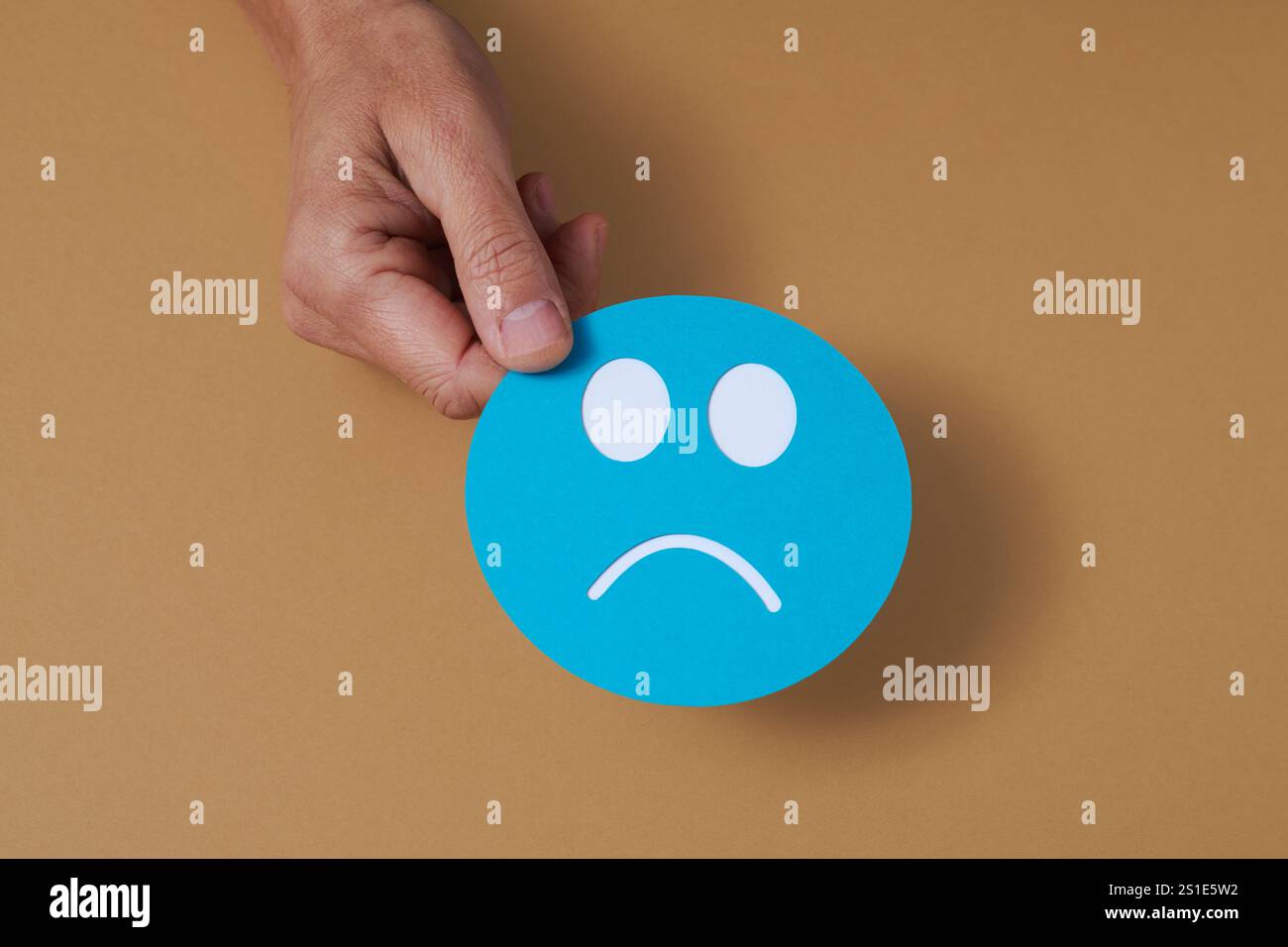 a man holds a blue paper sad face in his hand on a brown background ...
