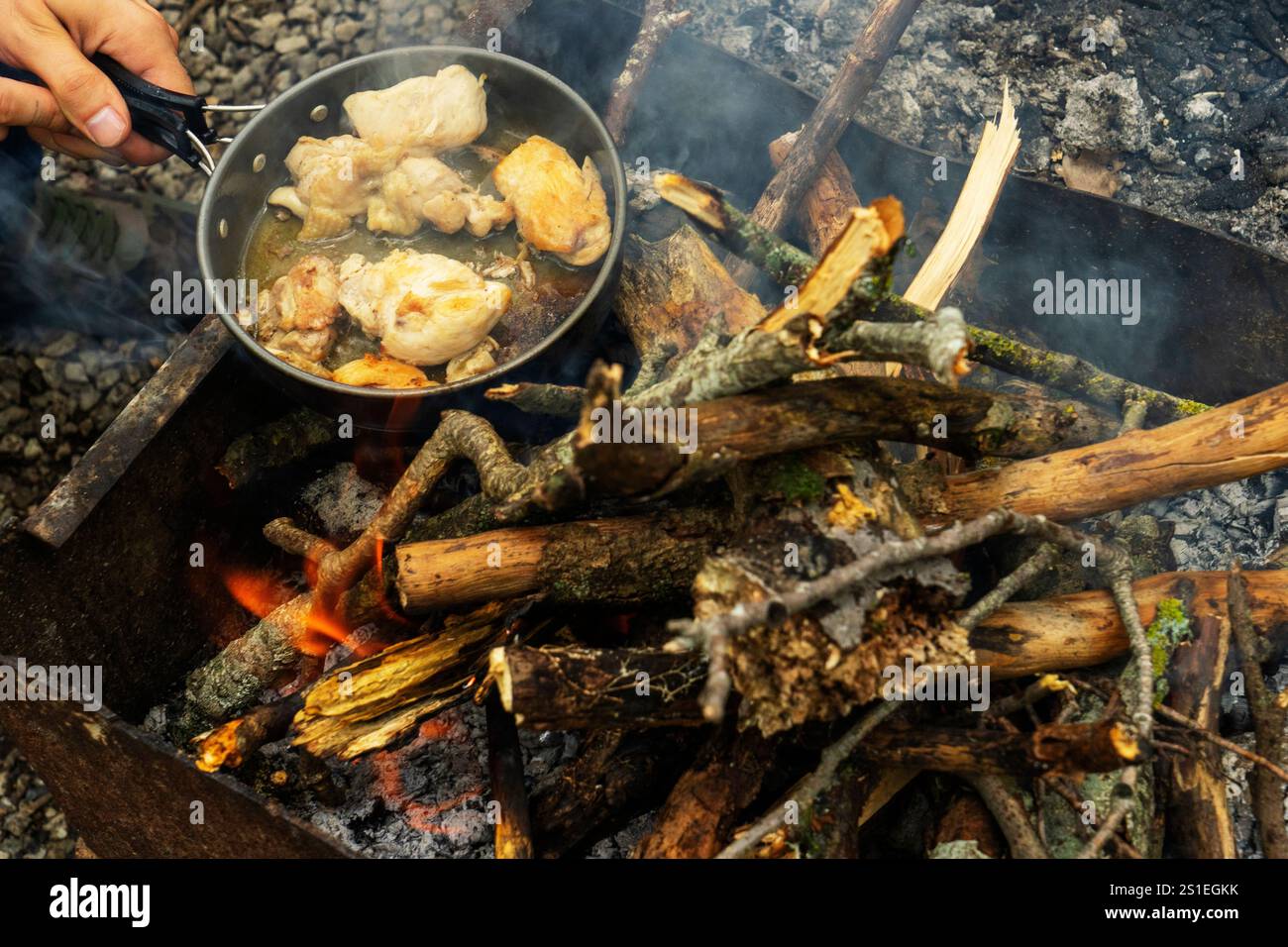 A cast-iron frying pan with chicken meat stands over a fire on a metal ...