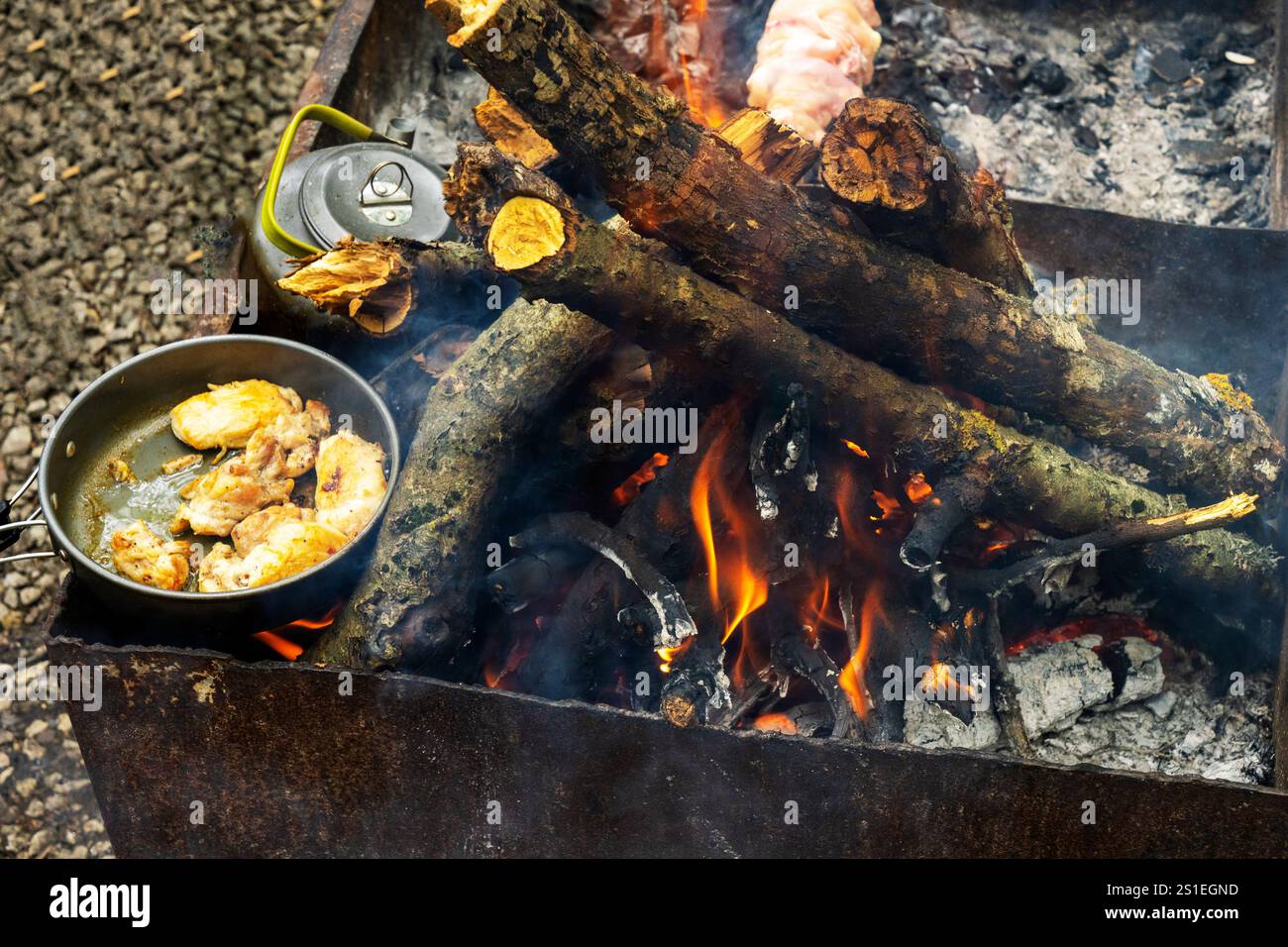 A cast-iron frying pan with chicken meat stands over a fire on a metal ...