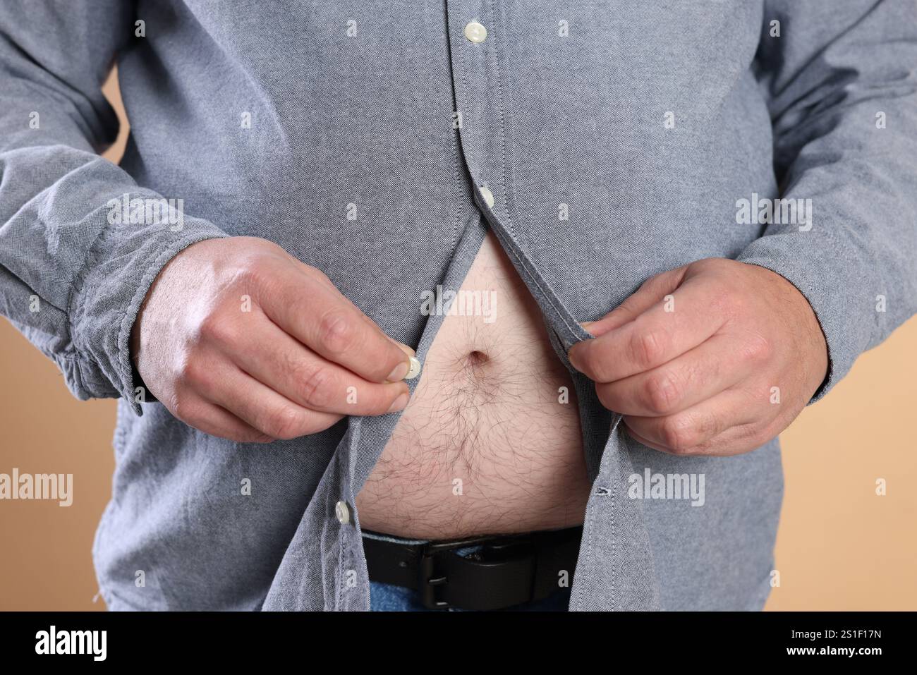 Overweight man trying to button up tight shirt on beige background ...