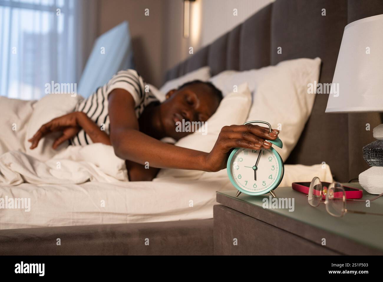 Young black woman lying in bed, turning off her alarm clock while ...