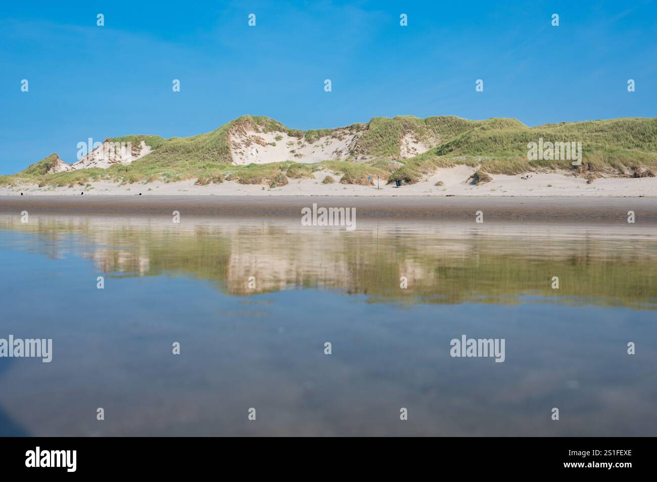 Sand dunes are reflected in the still, standing water at low tide at ...