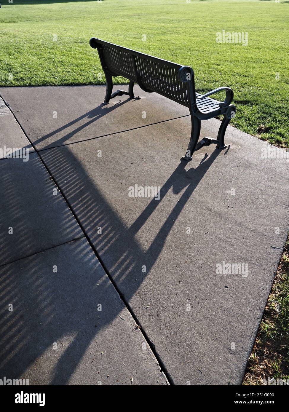 Park bench faces the rising sun in a city park. Shadows are long and ...