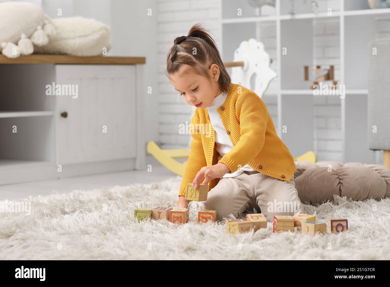 Cute little girl playing with wooden cubes and sitting on carpet at ...