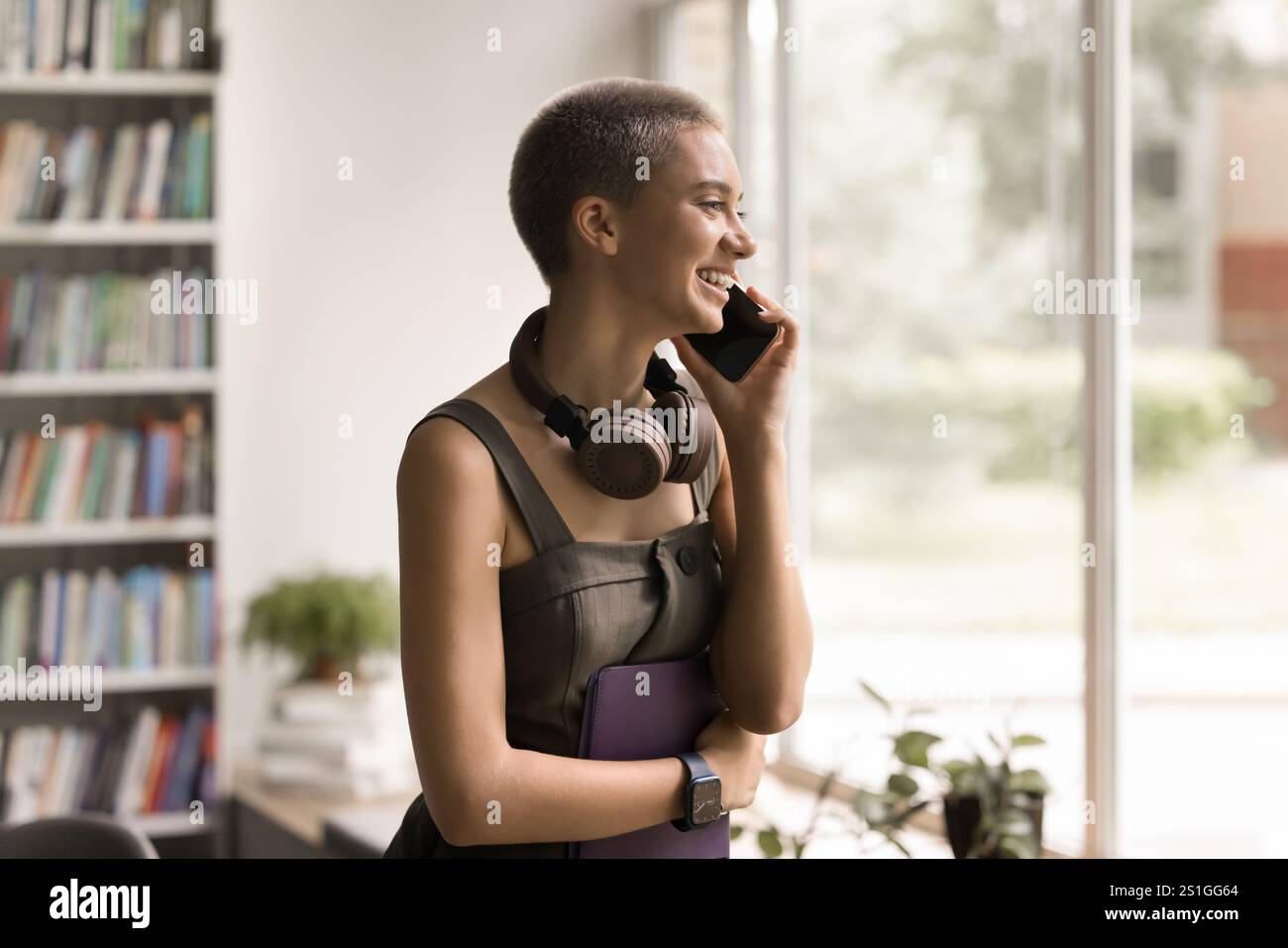 Smiling millennial female librarian stand by window calling by phone ...