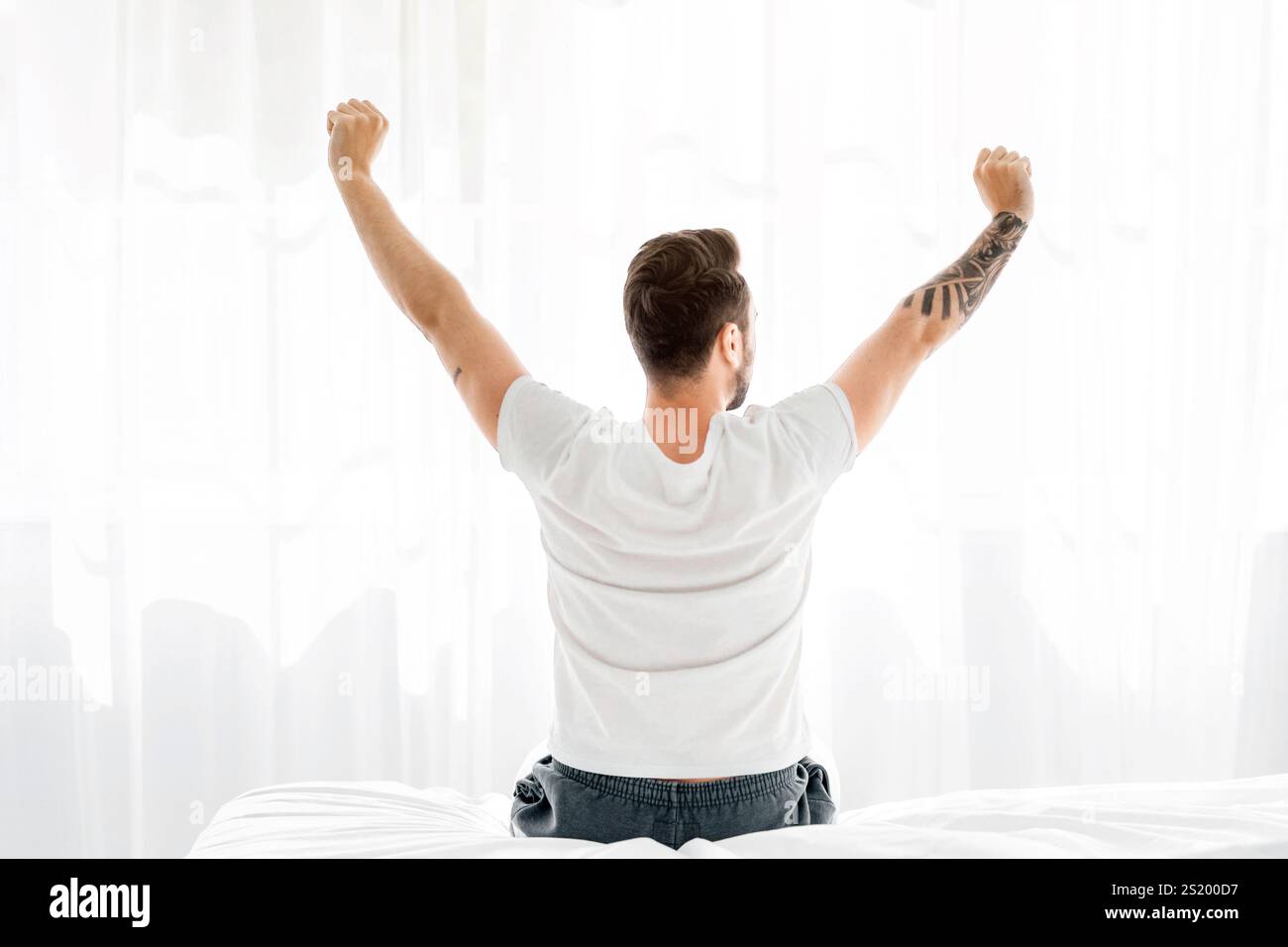 Relaxed young man waking up in bed and stretching his arms, facing the window, back view, free ...
