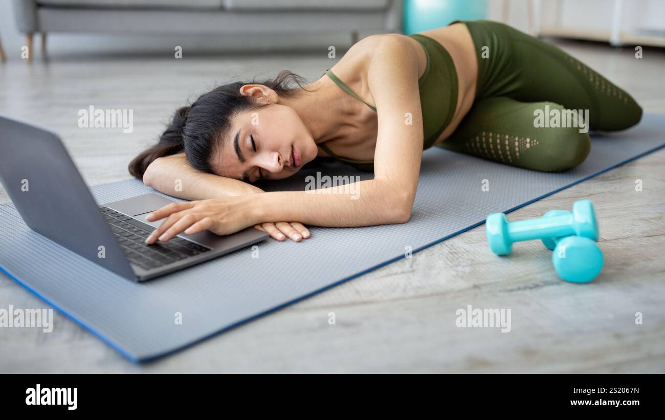 Tired young Indian woman sleeping on yoga mat in front of laptop, too ...