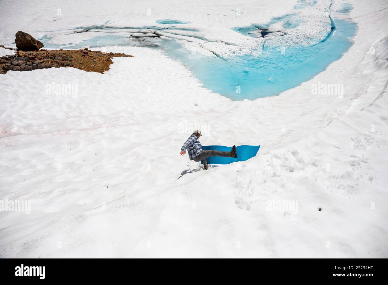 A man stands on a sleeping pad to slide down a hill to a frozen lake ...