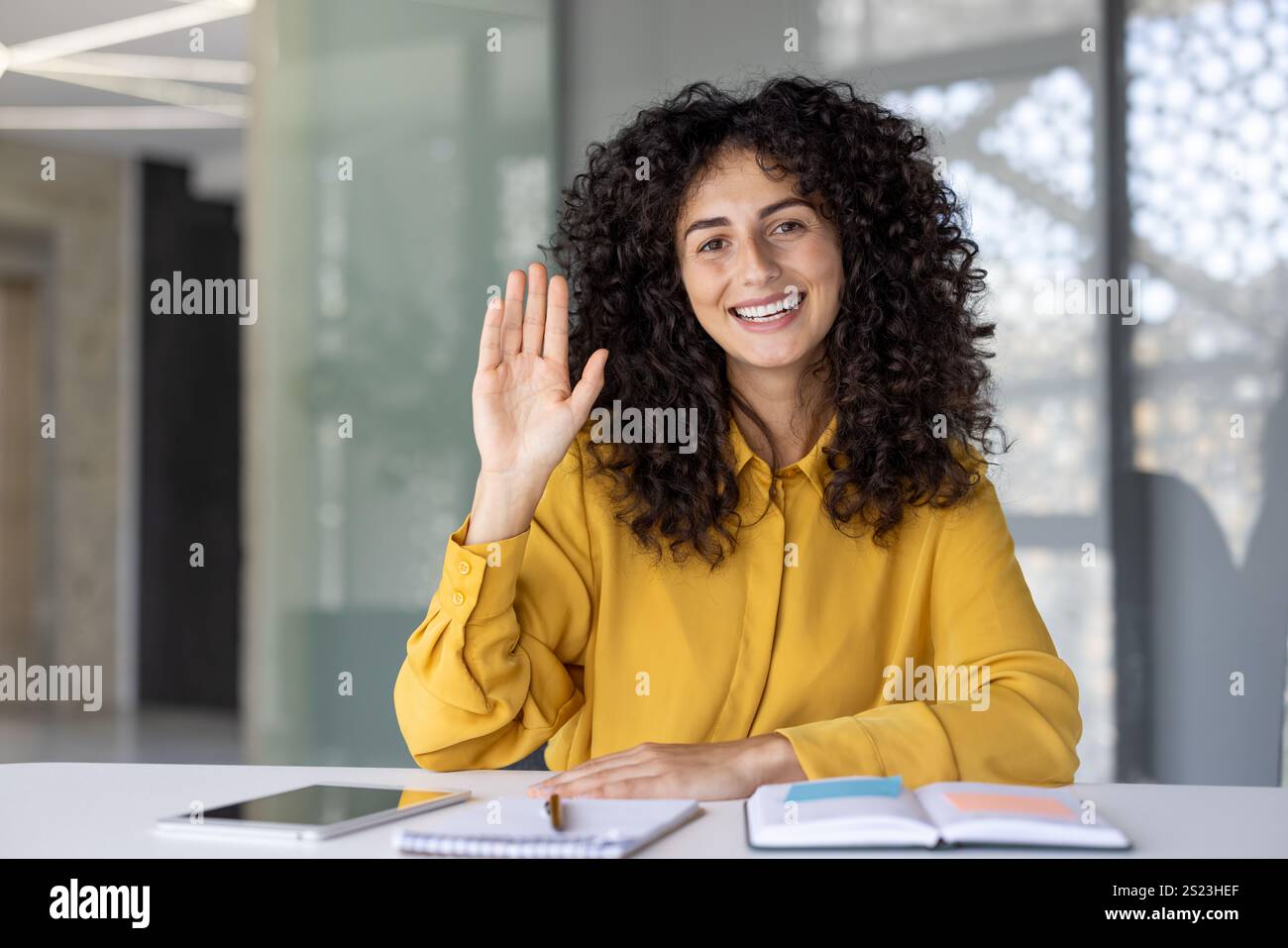 Woman sitting inside office at workplace looking at camera, waving ...