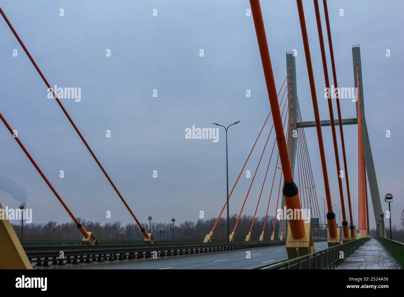 A modern suspension bridge features striking orange cables and a wide roadway under an overcast ...
