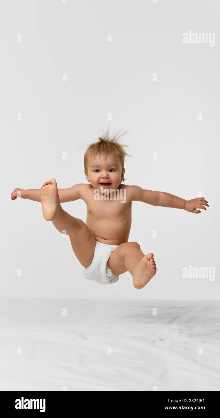A lively baby girl wearing a diaper jumps playfully with arms and legs ...