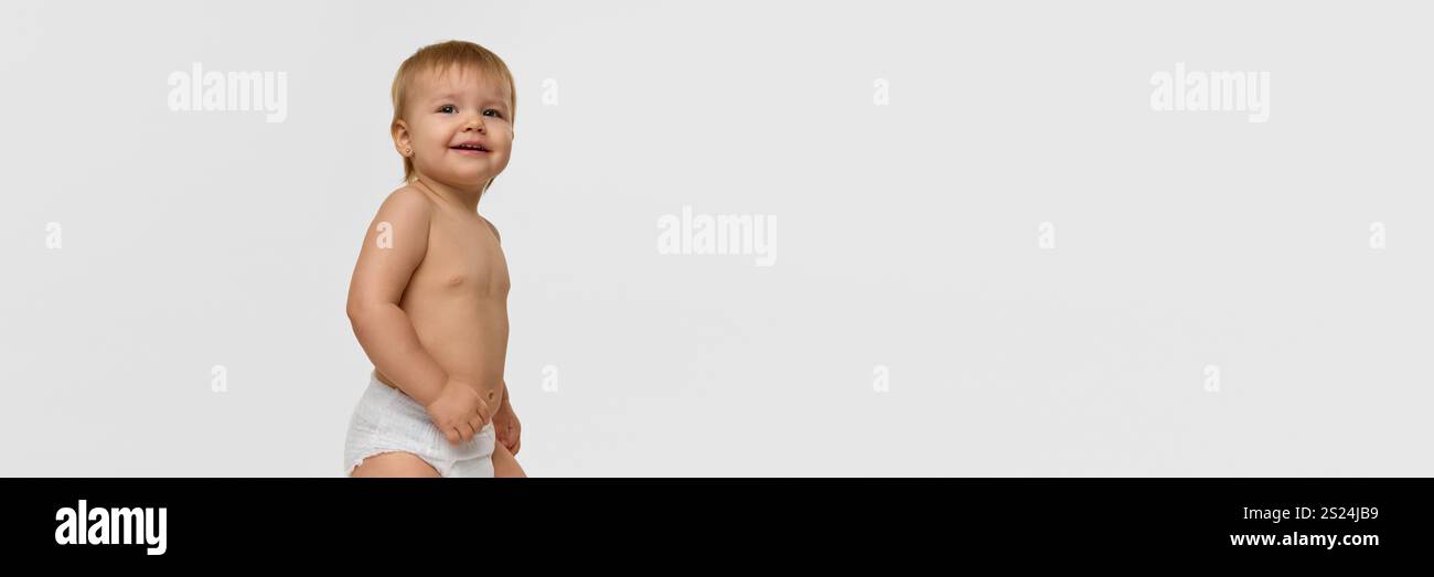 Cheerful baby girl in a diaper stands with a smile against studio ...