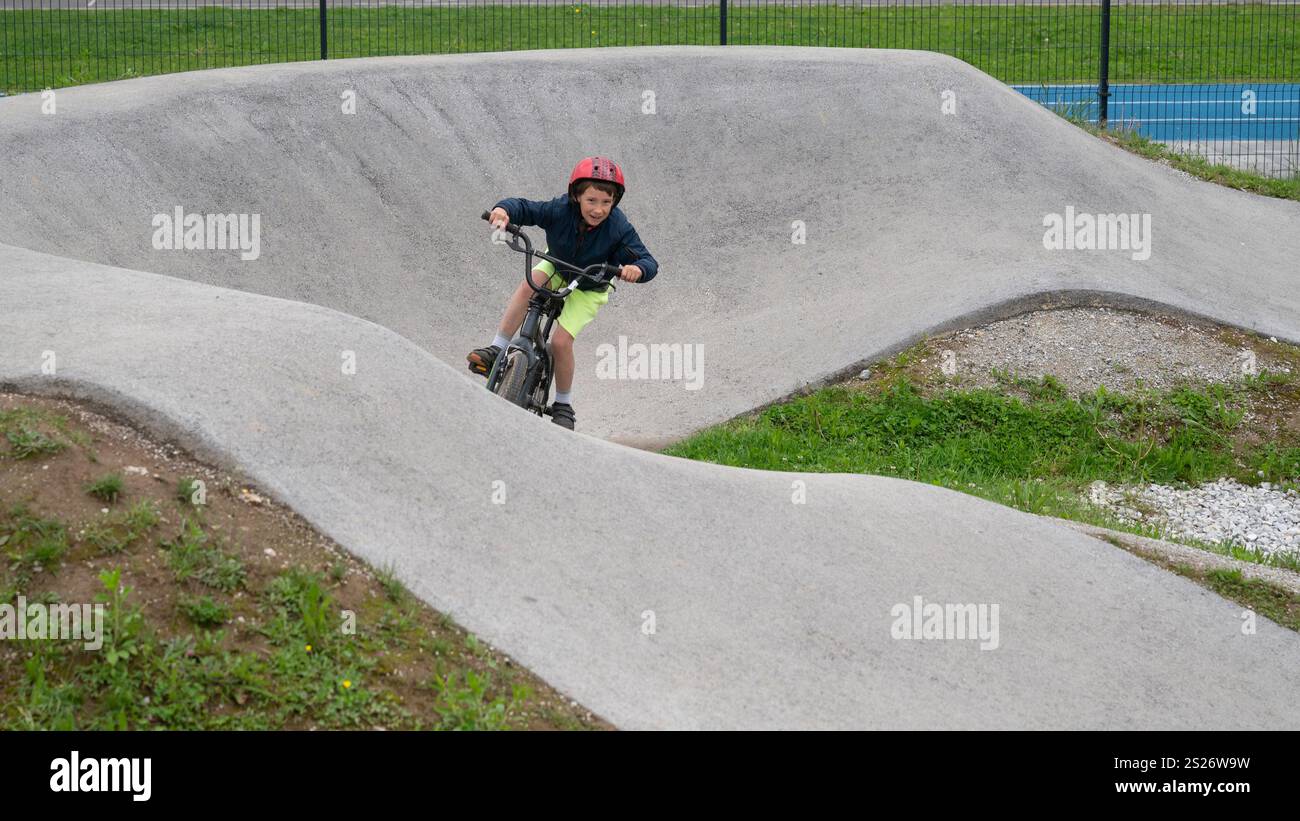 A child with a red helmet rides a bike enthusiastically on a pump track ...