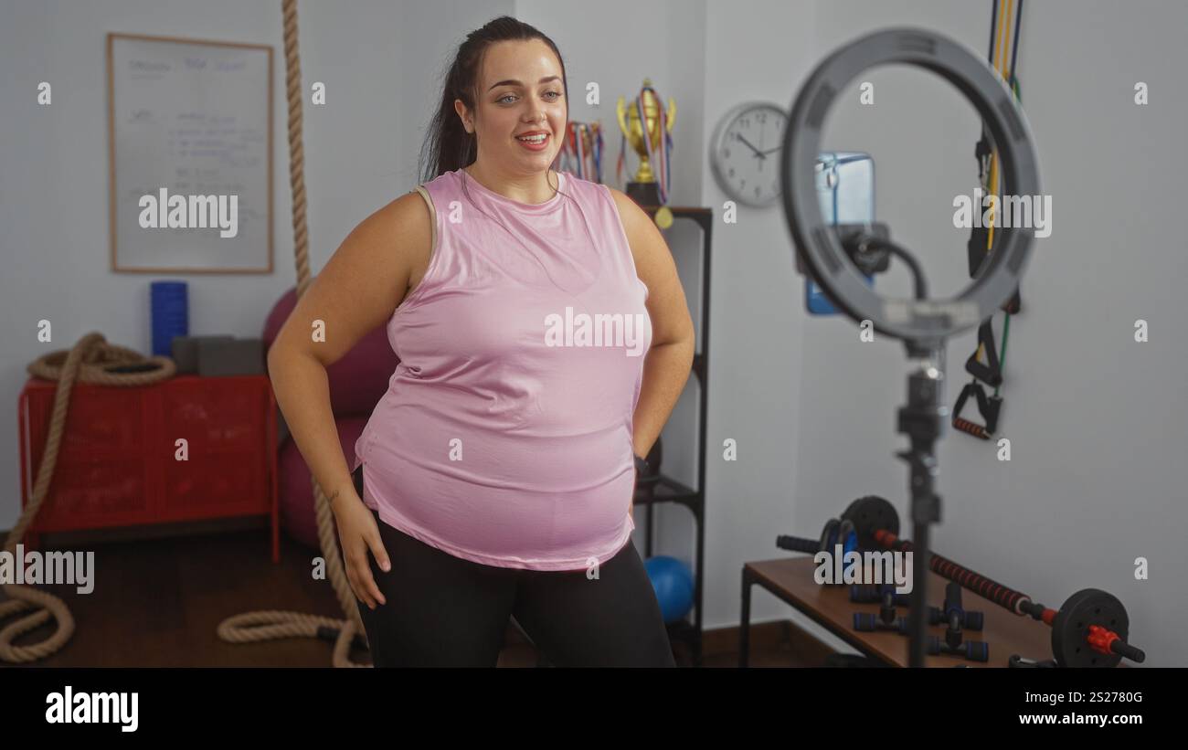 Woman filming content in gym wearing pink top standing next to sports ...