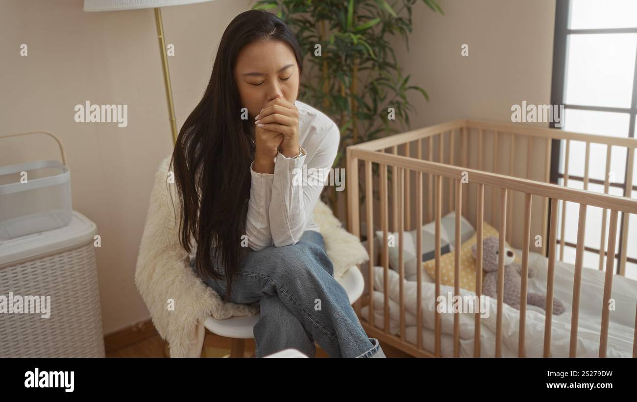 Woman sitting on chair in bedroom next to baby cradle, praying with ...
