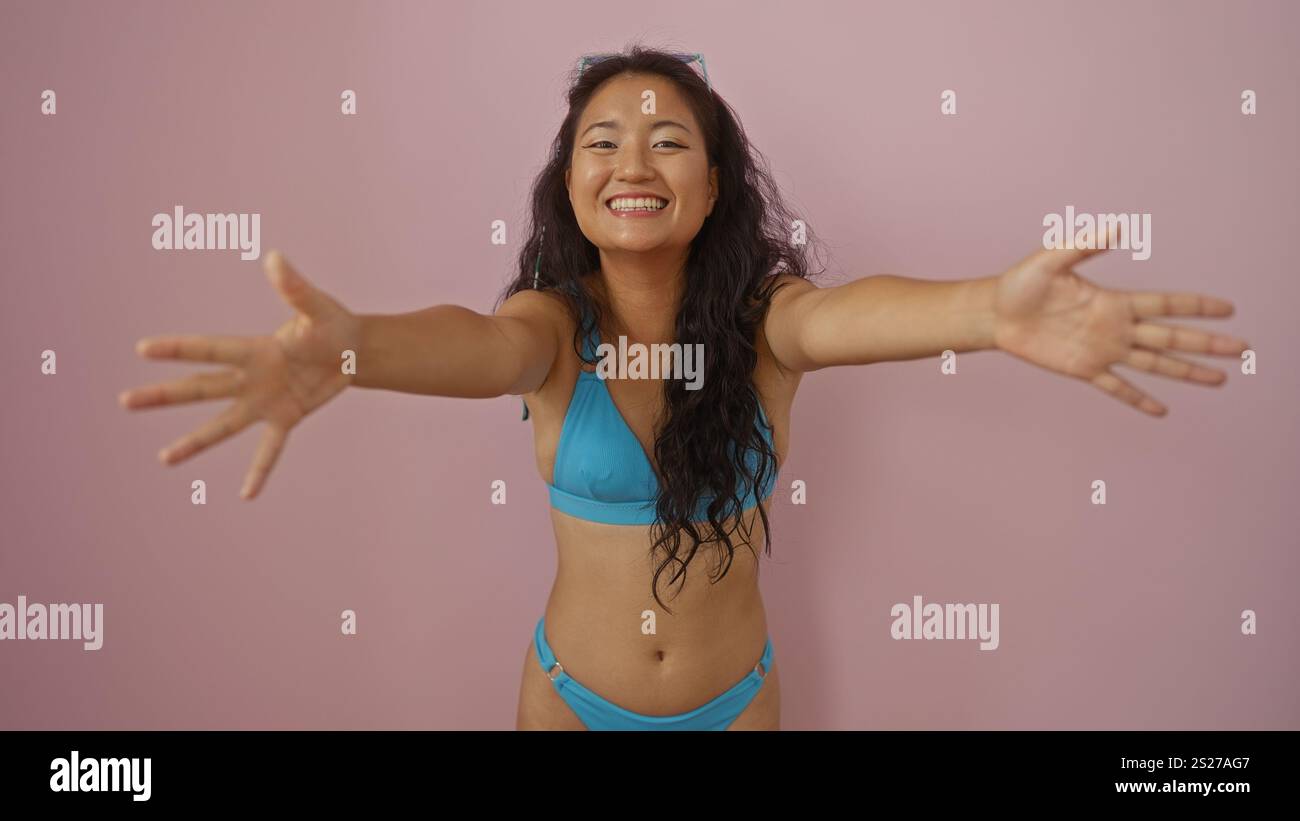 Young beautiful asian woman smiling in blue bikini against a pink wall, extending arms wide open ...