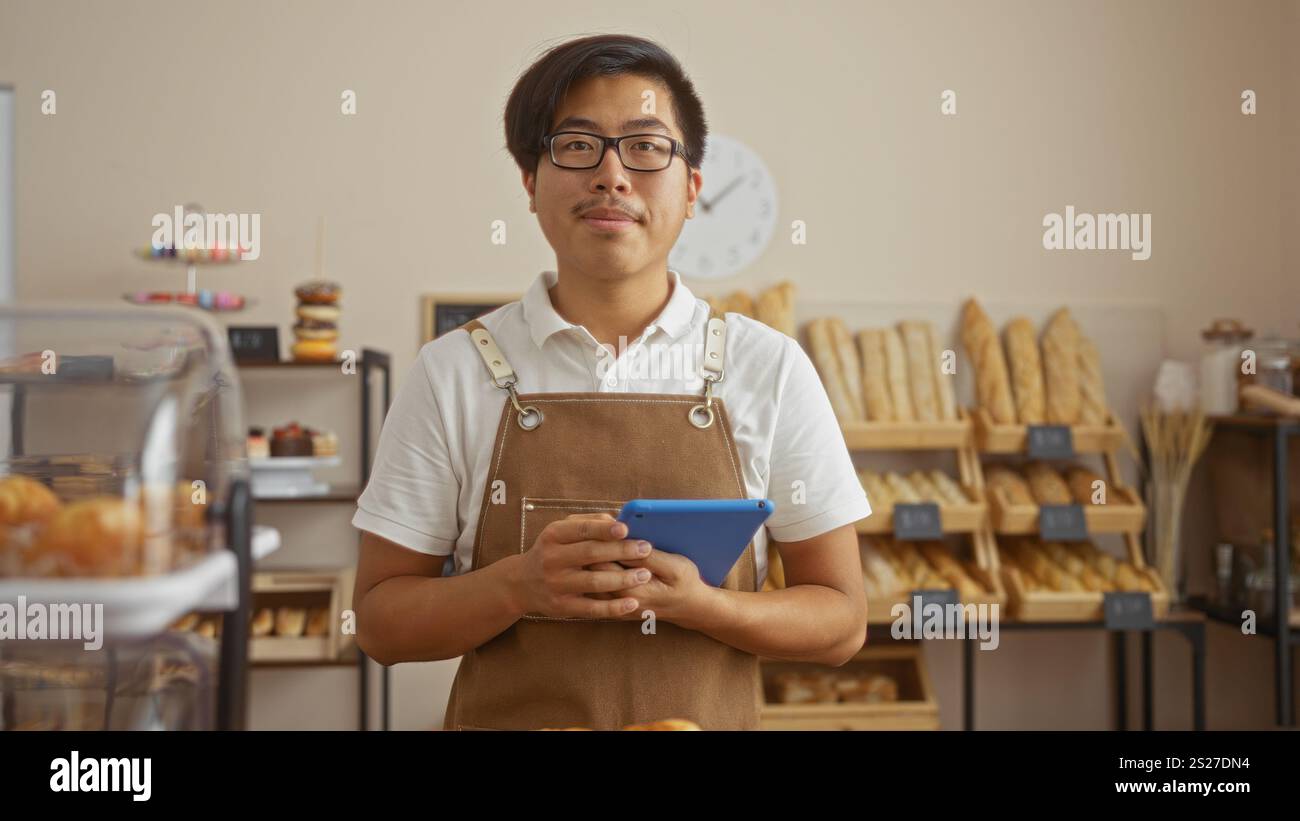 Young chinese man in bakery holding tablet surrounded by bread and ...