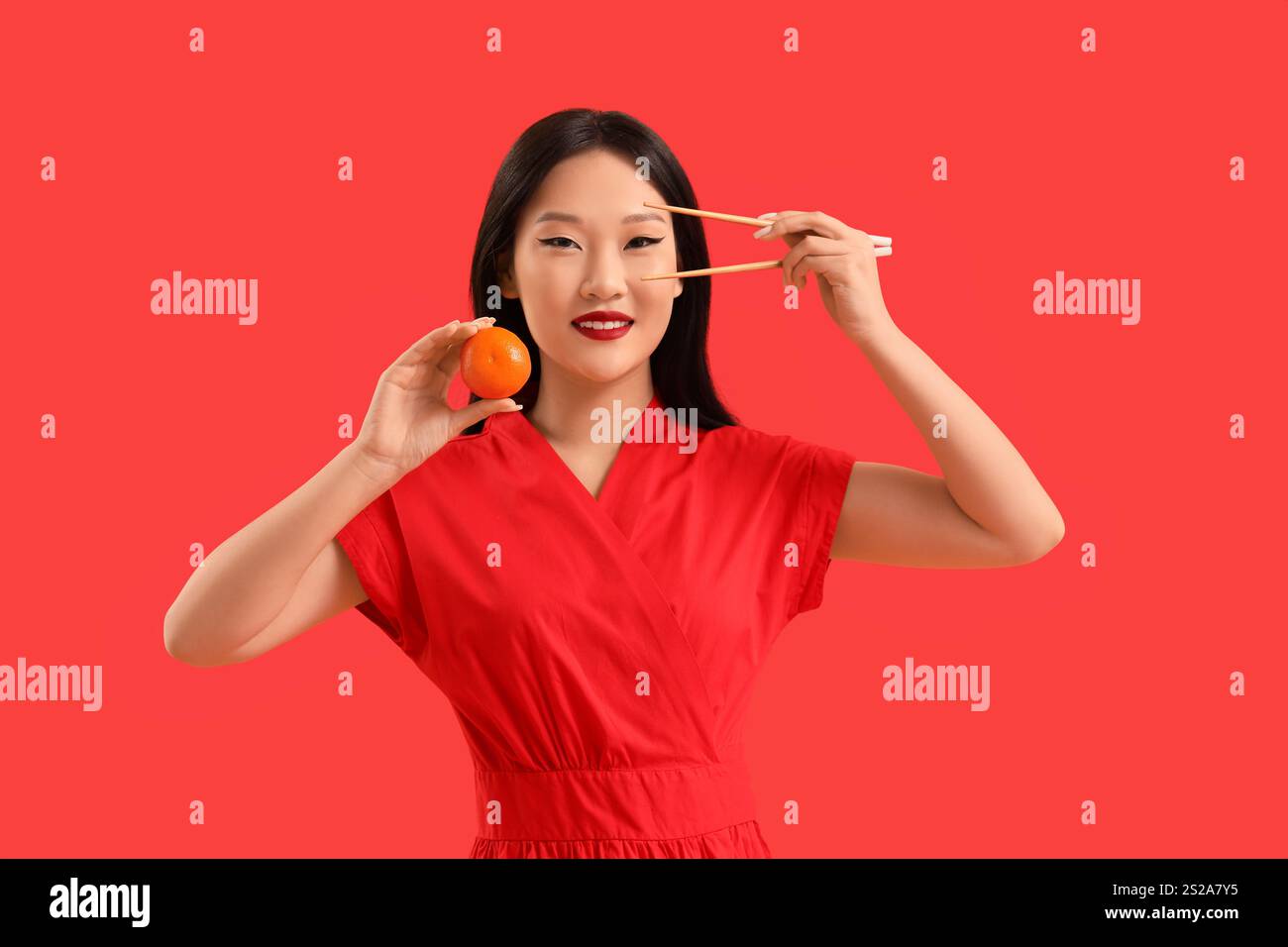Beautiful Asian woman with chopsticks and tangerine on red background ...