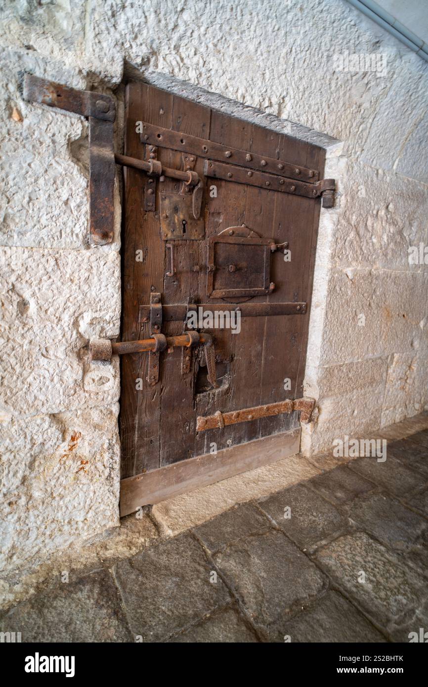 A weathered wooden prison door reinforced with iron bars, locks, and hinges, set within a thick ...