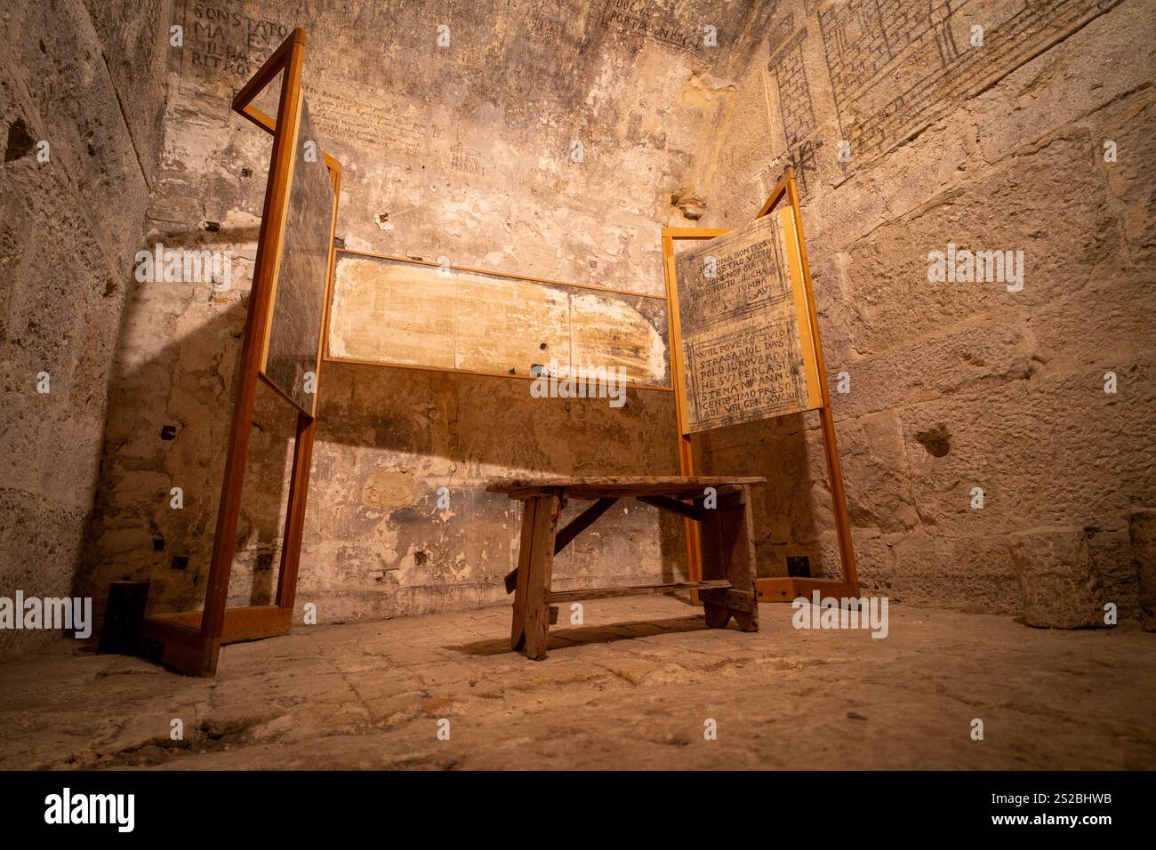A dimly lit stone cell featuring a rough wooden bench, walls covered in ...