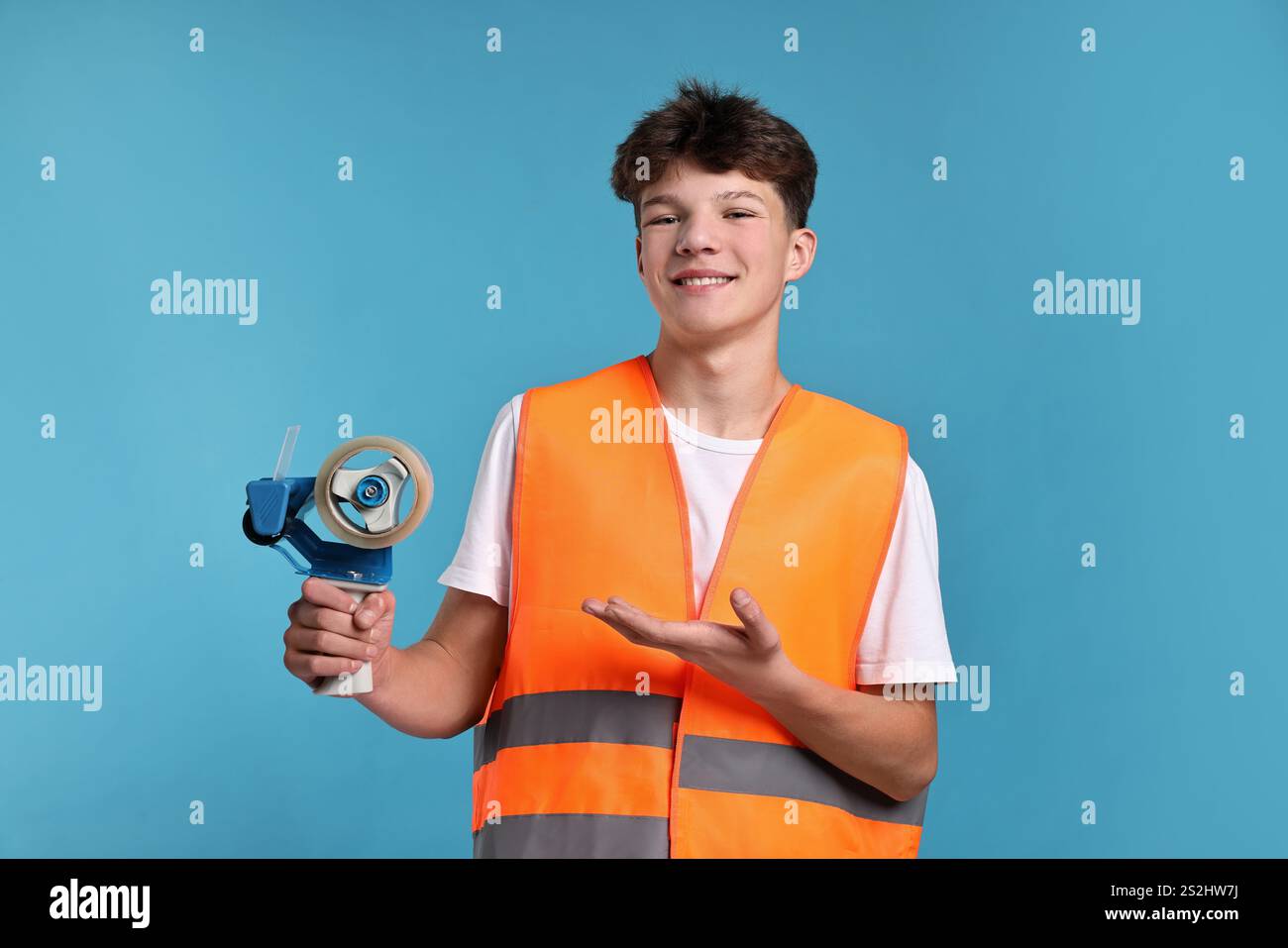 Teenage boy with tape gun dispenser in safety vest working as warehouse ...
