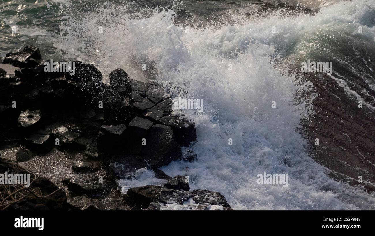 Powerful ocean waves crash against jagged volcanic rocks, creating a ...