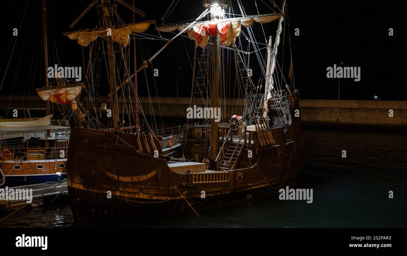 A historic-style wooden sailing ship docked at night, illuminated by a ...