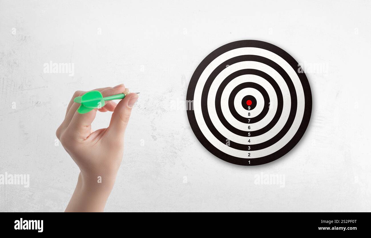 Hand holding a dart aiming at a black-and-white dartboard on a wall ...