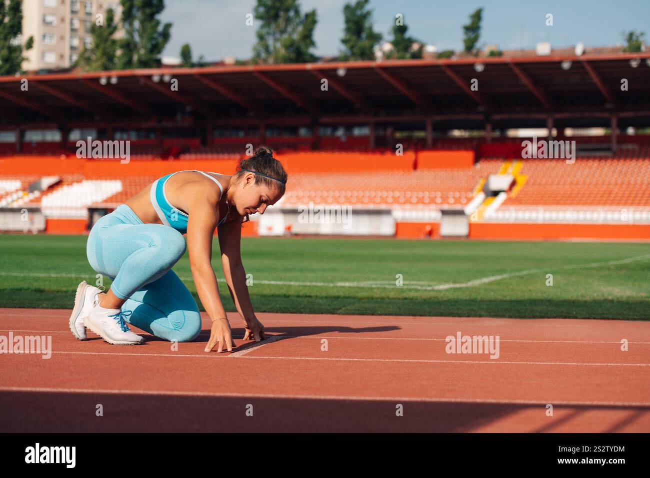 A fit female athlete is kneeling in start position on stadium. A ...