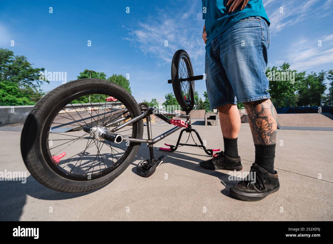 Legs of a tattooed man standing next to a broken bmx in a skate park ...