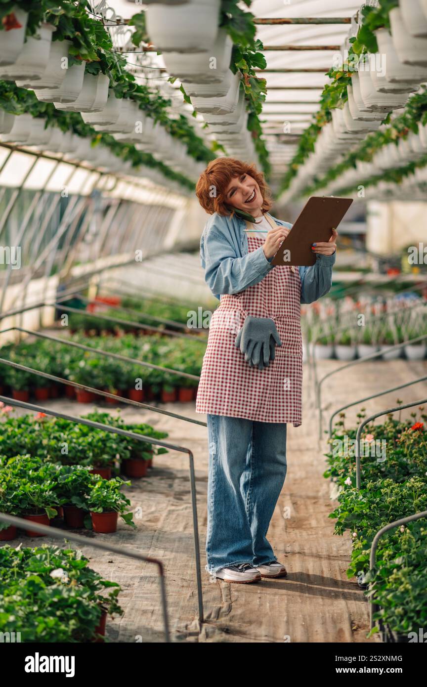 Full length portrait of cheerful female botanist standing at greenhouse with clipboard in hands ...
