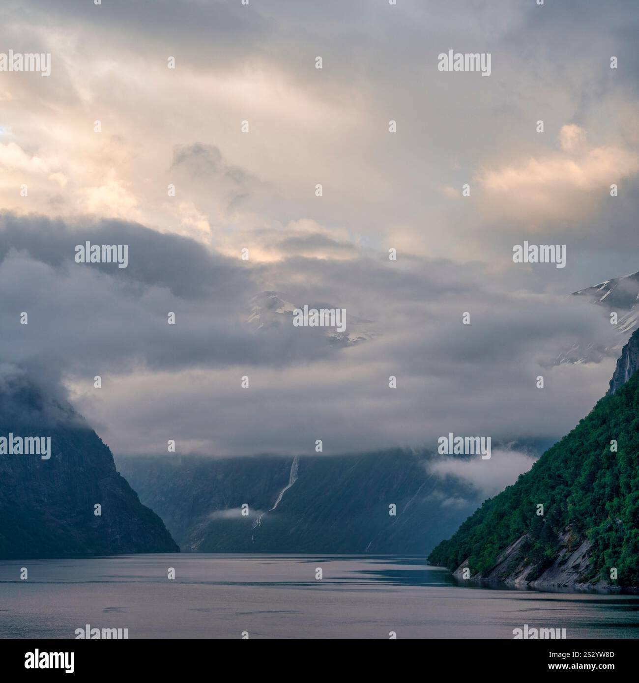 A calm fjord in Norway, surrounded by towering mountains shrouded in ...