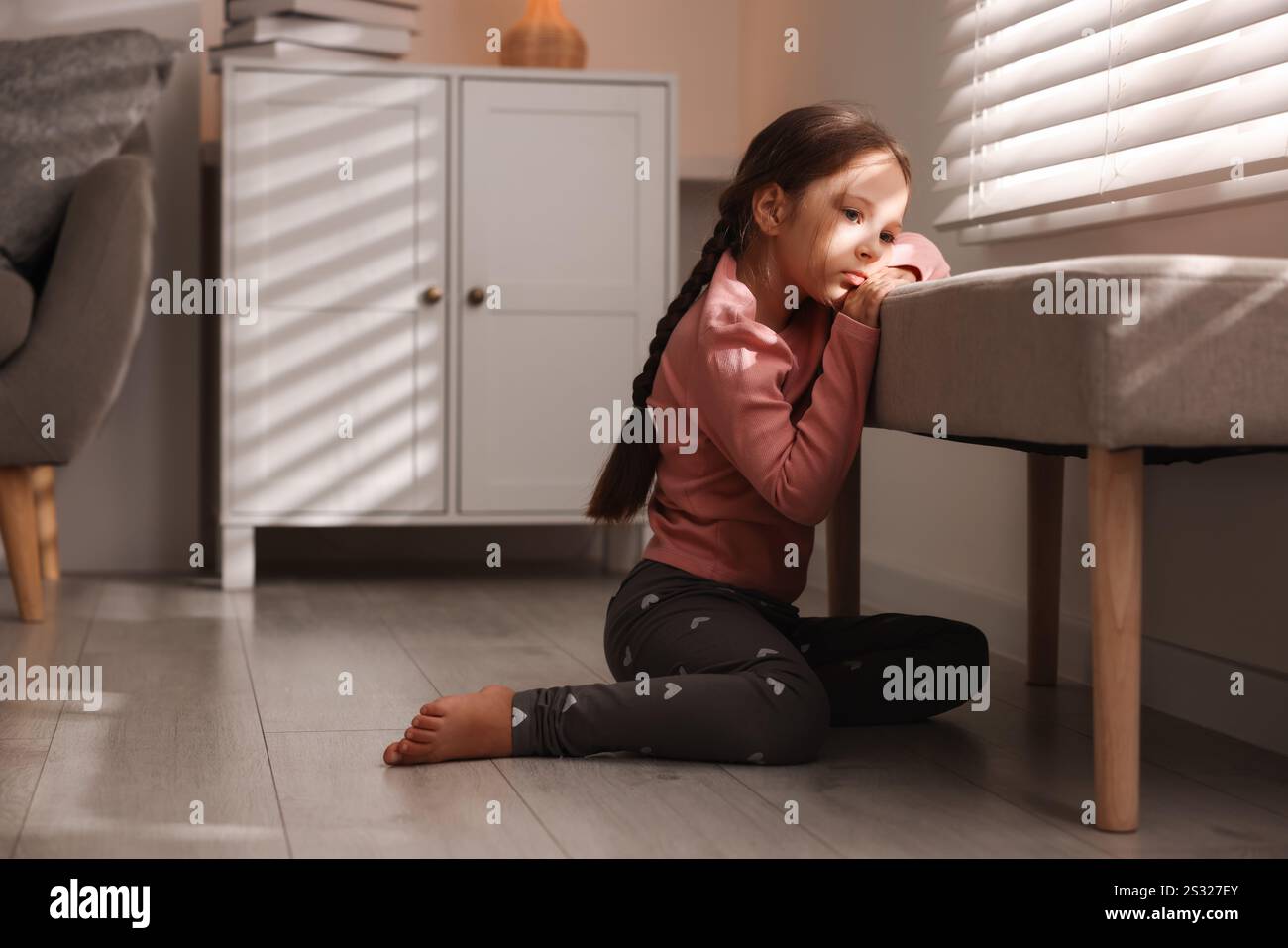 Orphanage concept. Sad girl and looking through window blinds indoors ...