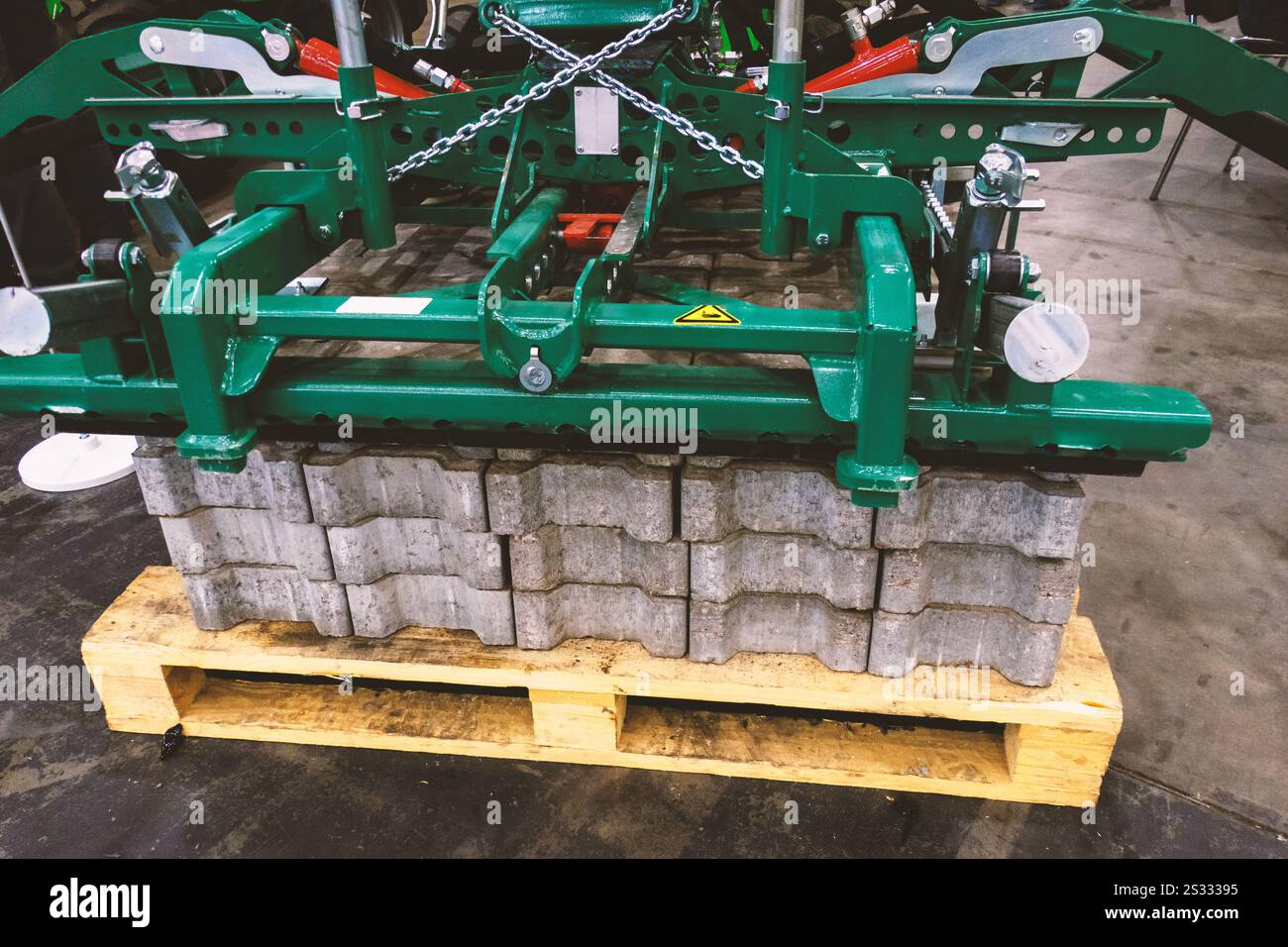 A green mechanical grab lifting a pallet of gray concrete paving stones ...