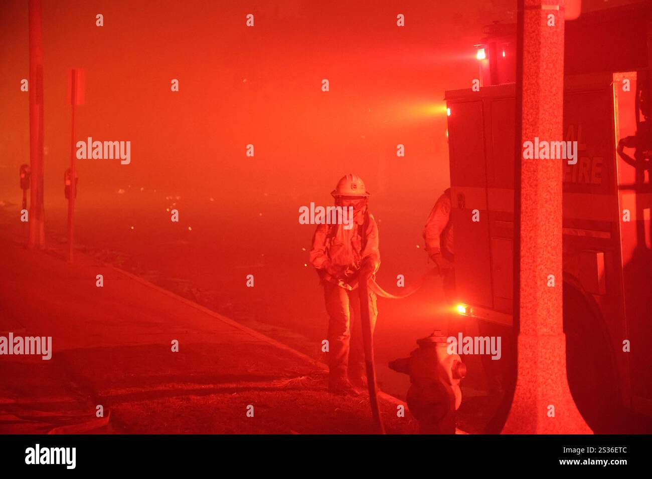 A firefighter manages a hose under the glow of nearby fires ...