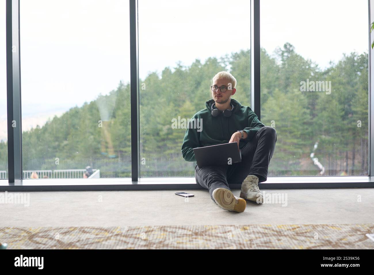 A blonde man sitting on the floor of a modern office, leaning against ...