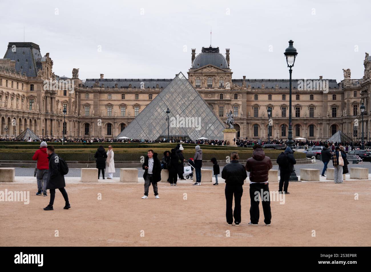 Crowds gather at the Louvre Museum under a gray sky with the iconic ...