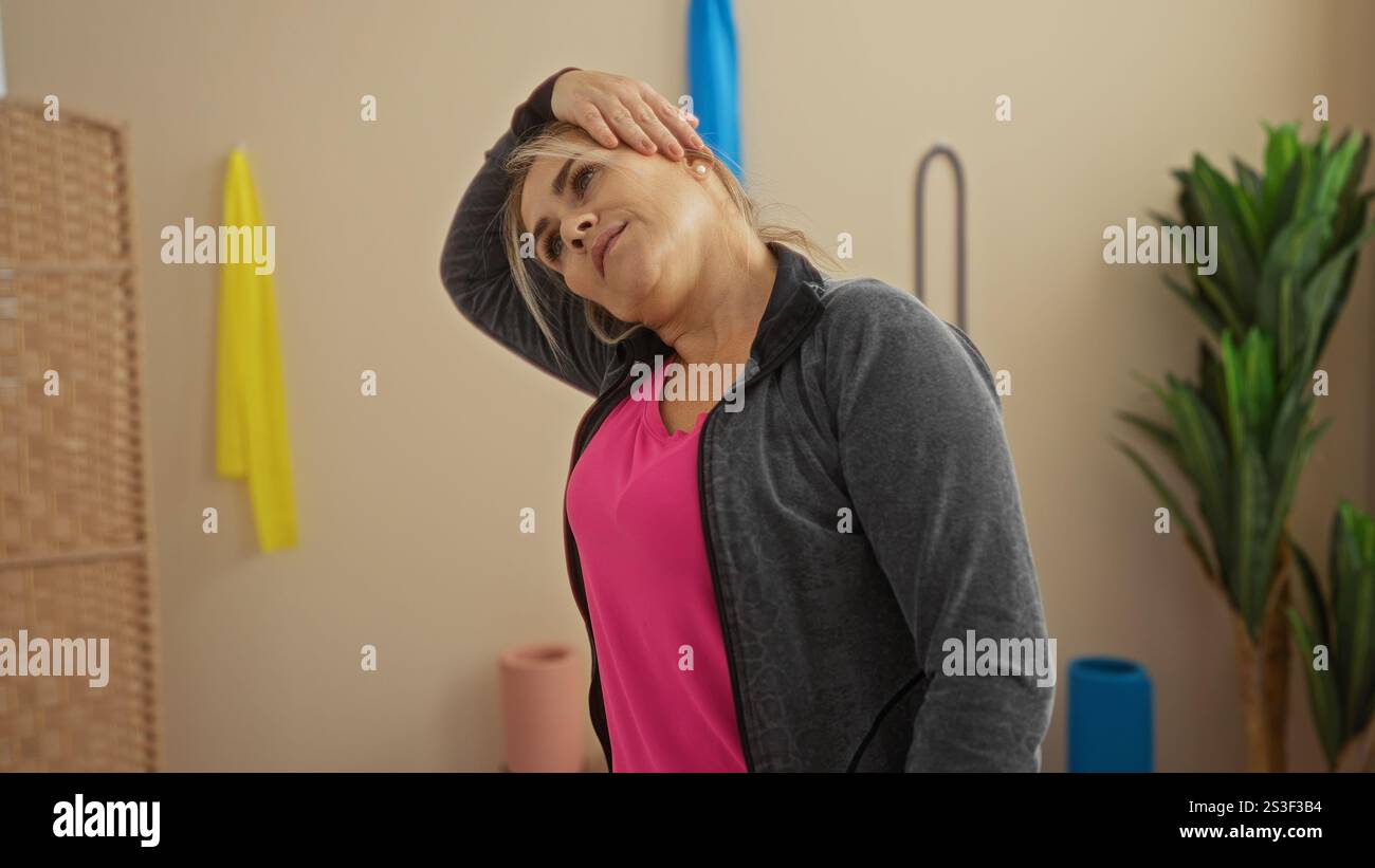 Woman stretching in gym, wearing pink shirt, blonde hair, grey jacket ...