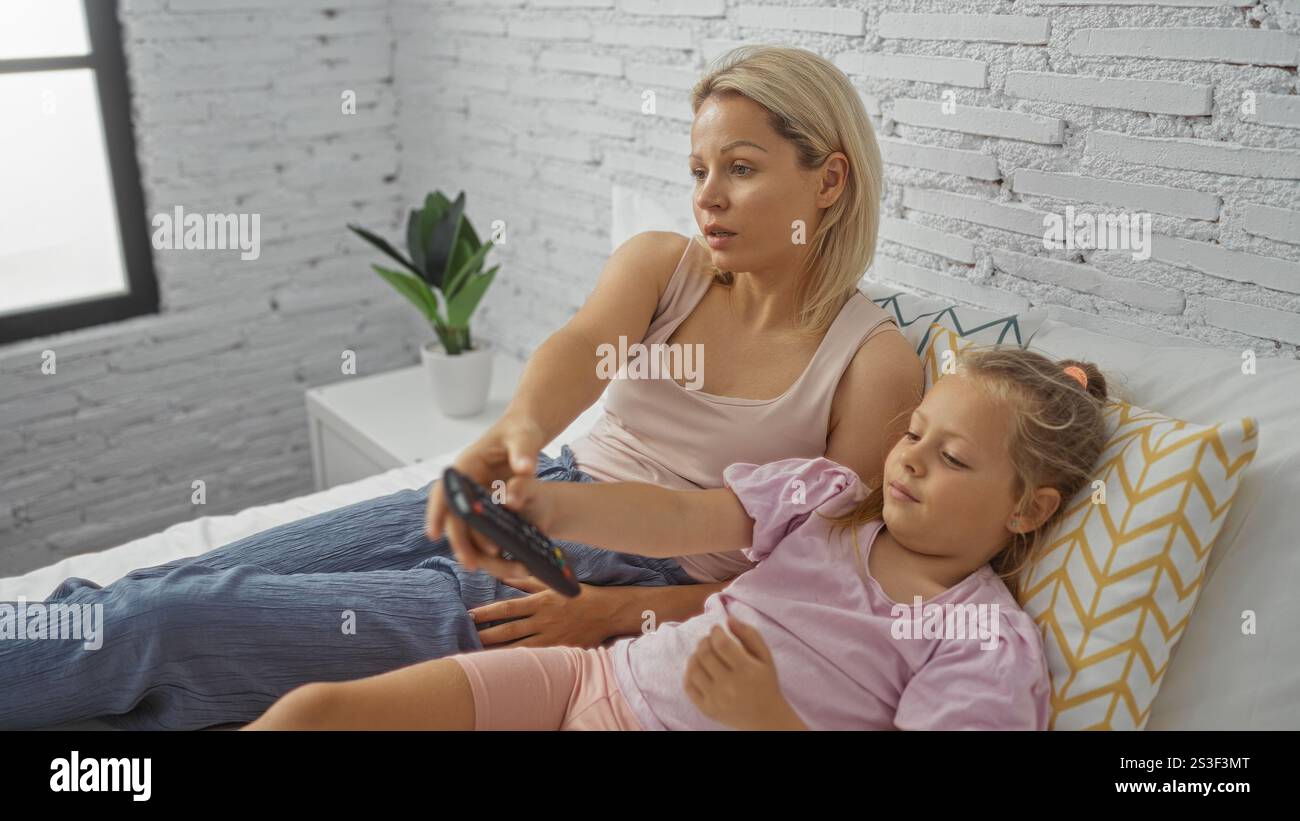 Woman watching tv with her daughter indoors on a bed in a modern home ...