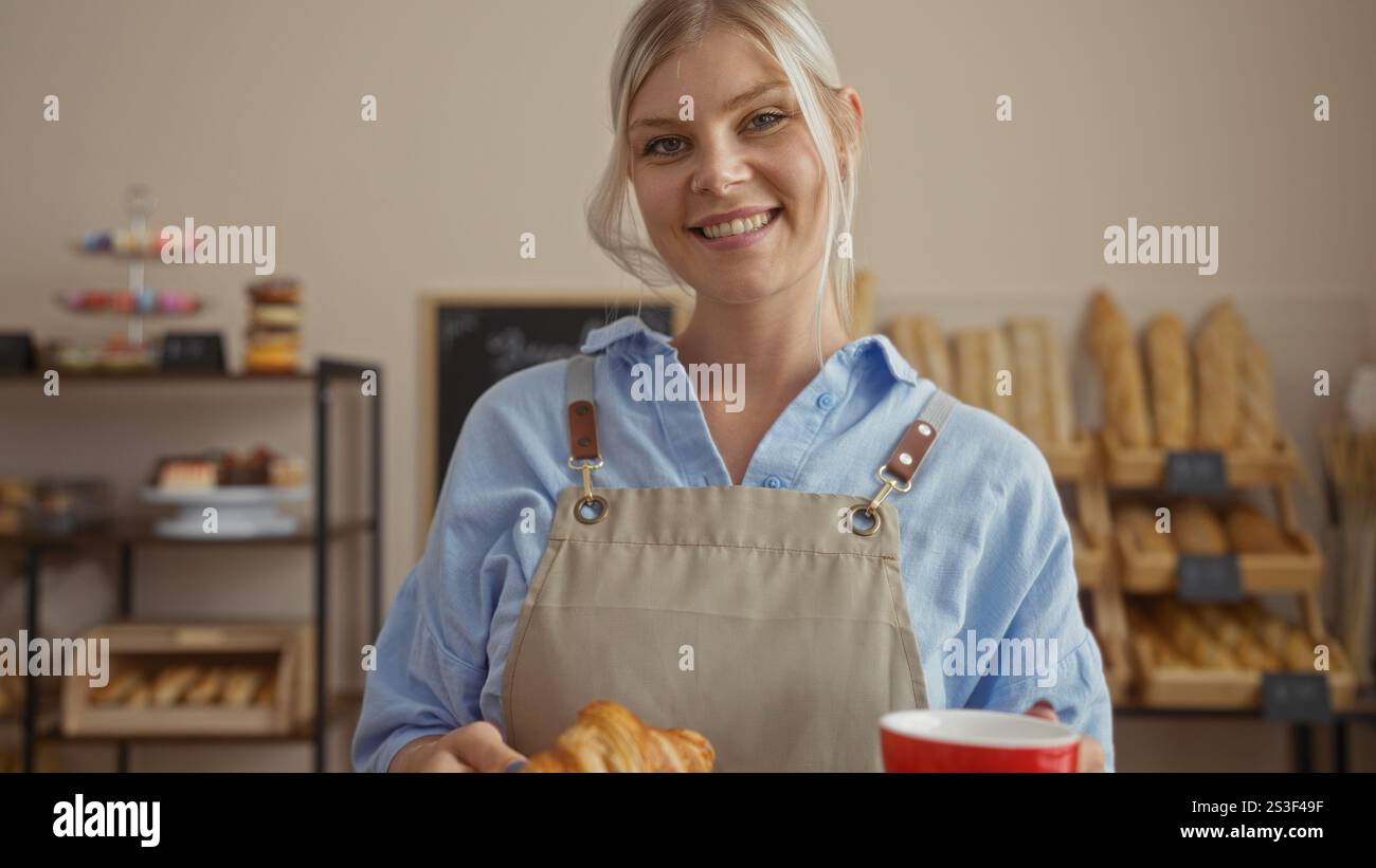 Young blonde woman in a bakery holding a croissant and a coffee cup ...