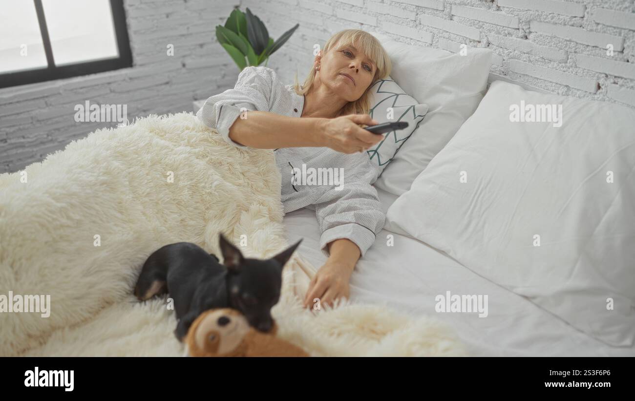Middle-aged woman lounging in bed at home with blonde hair, watching television while a ...
