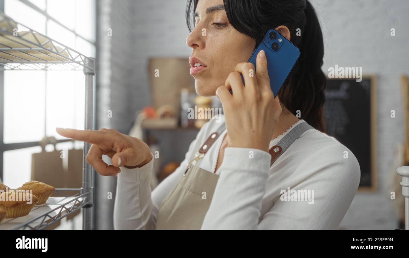 Hispanic woman talking on phone in bakery while pointing at muffins on ...