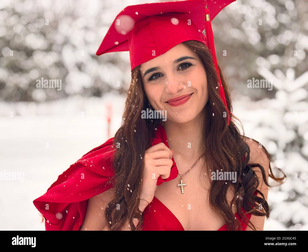 Graduating girl with long brunette hair wearing red cap, and graduation gown in snowy winter ...