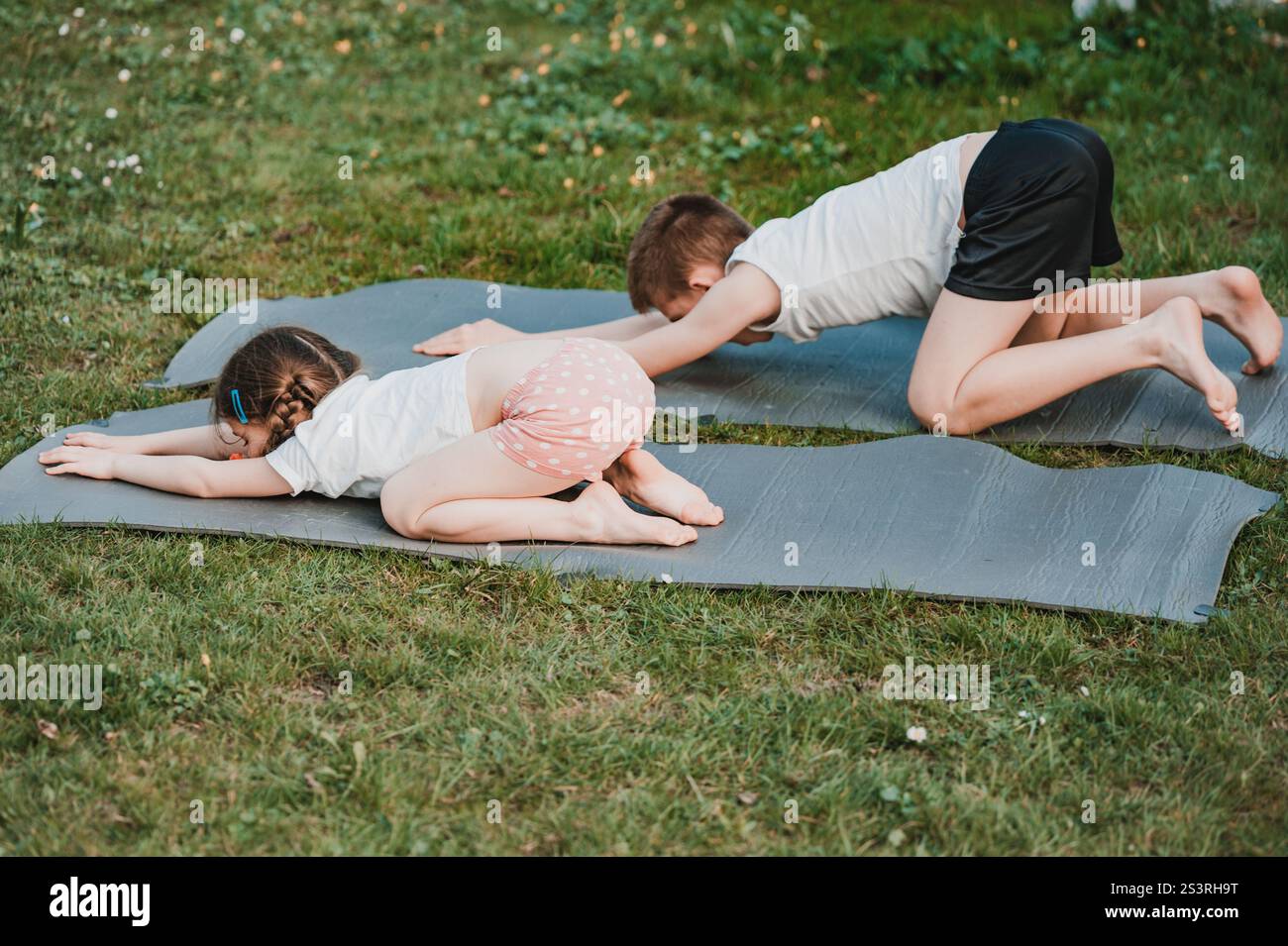 Two young children perform child's pose on yoga mats placed on a grassy lawn. They're enjoying a ...