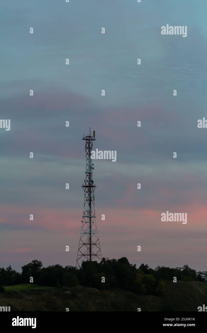 A tall communication tower rises above trees, silhouetted against a sunset sky with soft pink ...