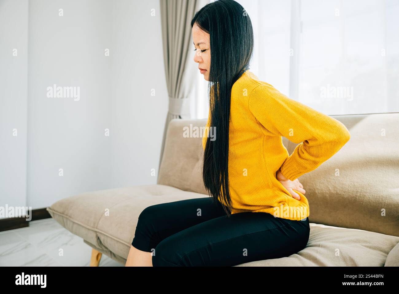 Asian woman holds her lower back in unbearable pain on a sofa Stock ...