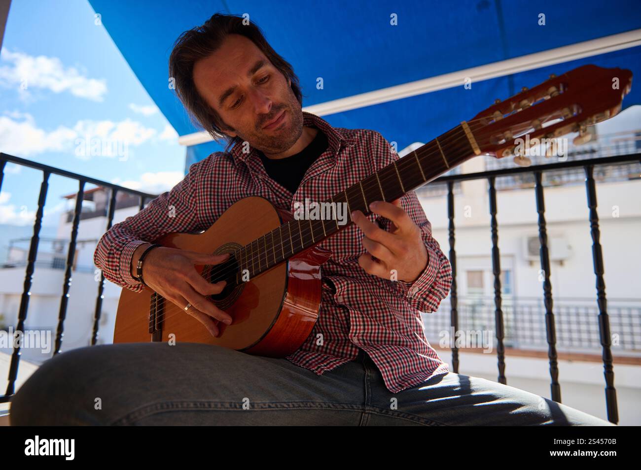 A person seated outdoors on a balcony, strumming an acoustic guitar ...