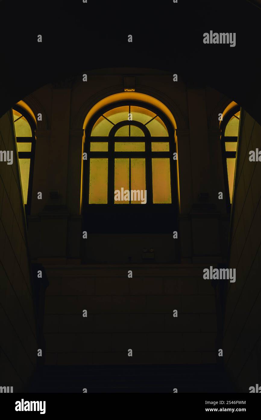 Low-light interior view of a staircase facing a tall arched window ...