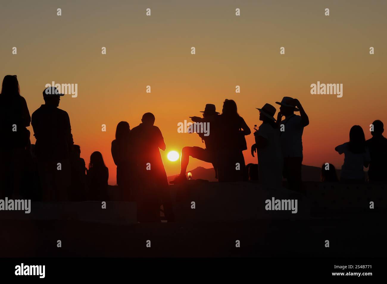 Silhouetted figures gather along a ledge to watch the sunset, their ...